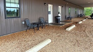 Uma área de estar coberta ao ar livre, com cadeiras e mesas, chão de cascalho e um edifício com fachada metálica.