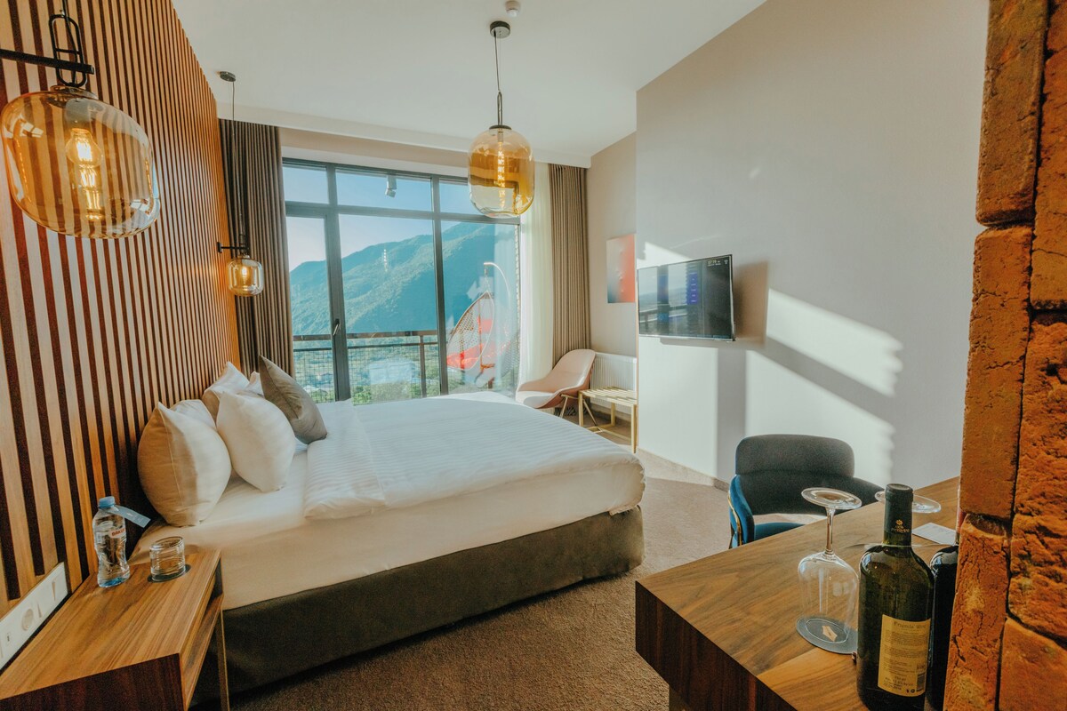 Quarto de hotel moderno com uma cama grande, uma mesa de cabeceira de madeira, um espaço de estar com uma cadeira, uma TV fixada na parede e vista para montanhas pela janela.