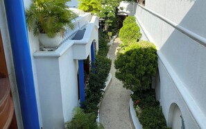 Um caminho estreito ladeado por paredes brancas e detalhes azuis, que conduz a uma área coberta com vista para o verde.