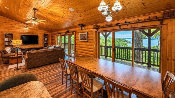 Interior de uma cabana de tronco com sala de jantar, sofá, televisão e vista para um lago.