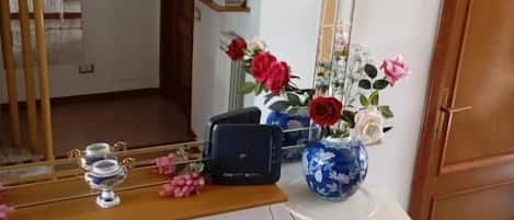 Quarto com uma escrivaninha de madeira, um espelho, um vaso com flores, uma pequena estante e uma porta.