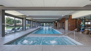 Piscina interna com paredes de vidro transparente, piso ladrilhado e vista para o exterior.