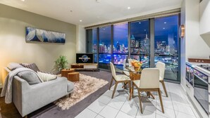 Uma sala de estar moderna com um sofá, uma televisão, uma mesa de jantar com cadeiras e vista para a cidade através de grandes janelas.