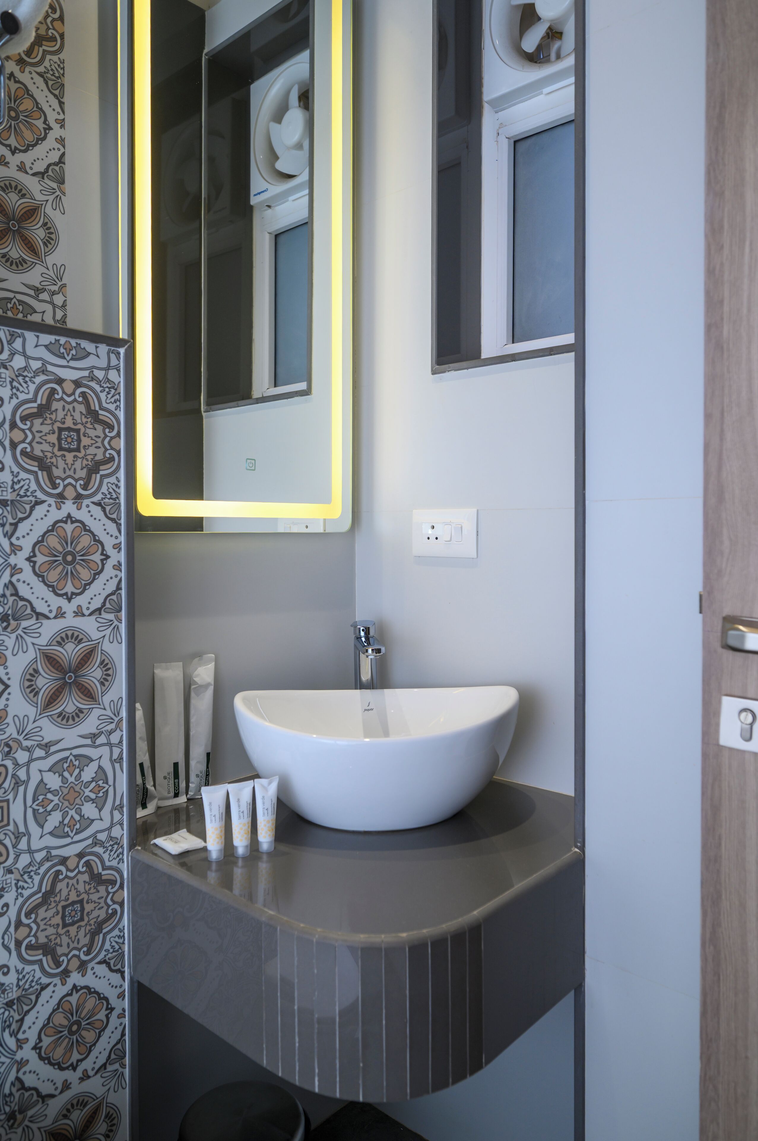 Um banheiro com pia branca, espelho iluminado e azulejos estampados.