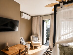 Quarto de hotel moderno com TV de tela plana, mesa de jantar de madeira, poltrona confortável e varanda com cortinas.