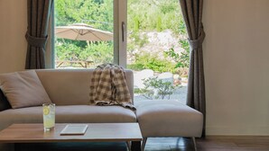 Uma sala de estar moderna com um sofá, uma mesa com um tablet e um copo de água, e vista para um jardim através de grandes janelas.