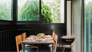 Uma sala de jantar com uma mesa posta para quatro pessoas, uma churrasqueira com carne e vista para o verde através de grandes janelas.