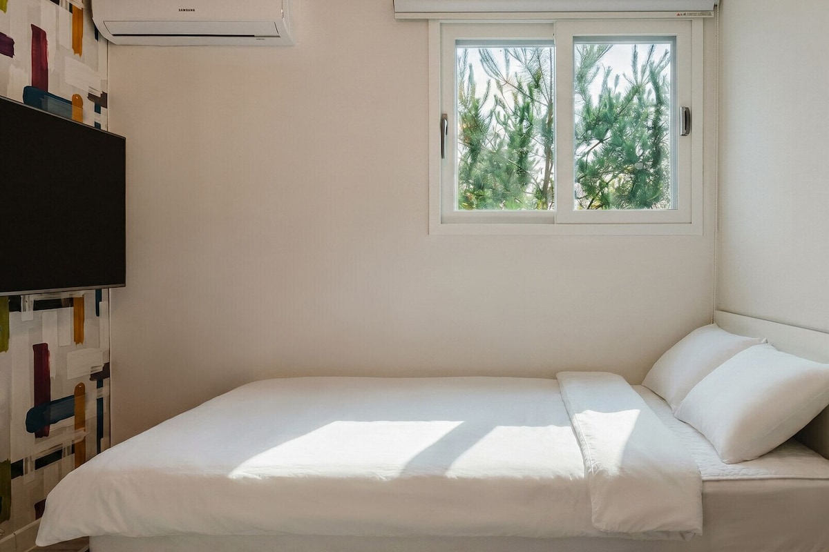 Uma cama de solteiro com roupa de cama branca, uma janela com vista para árvores e um ar condicionado fixado na parede.