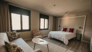 Quarto de hotel com uma cama, um sofá, uma cadeira, uma mesa pequena e duas janelas.