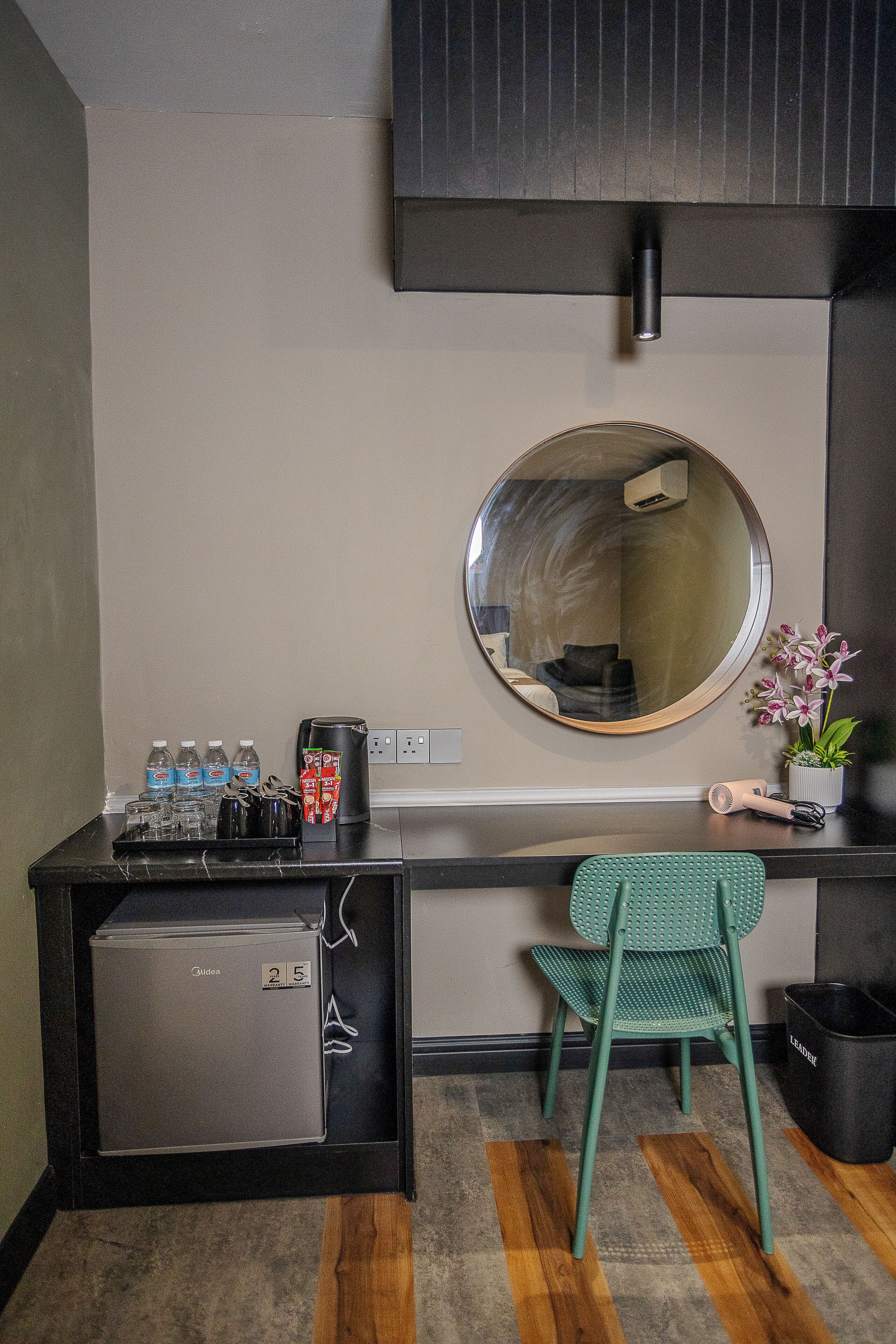 Quarto de hotel compacto com cozinha compacta, um espelho redondo, um pequeno frigorífico, uma cadeira verde e uma planta em vaso.