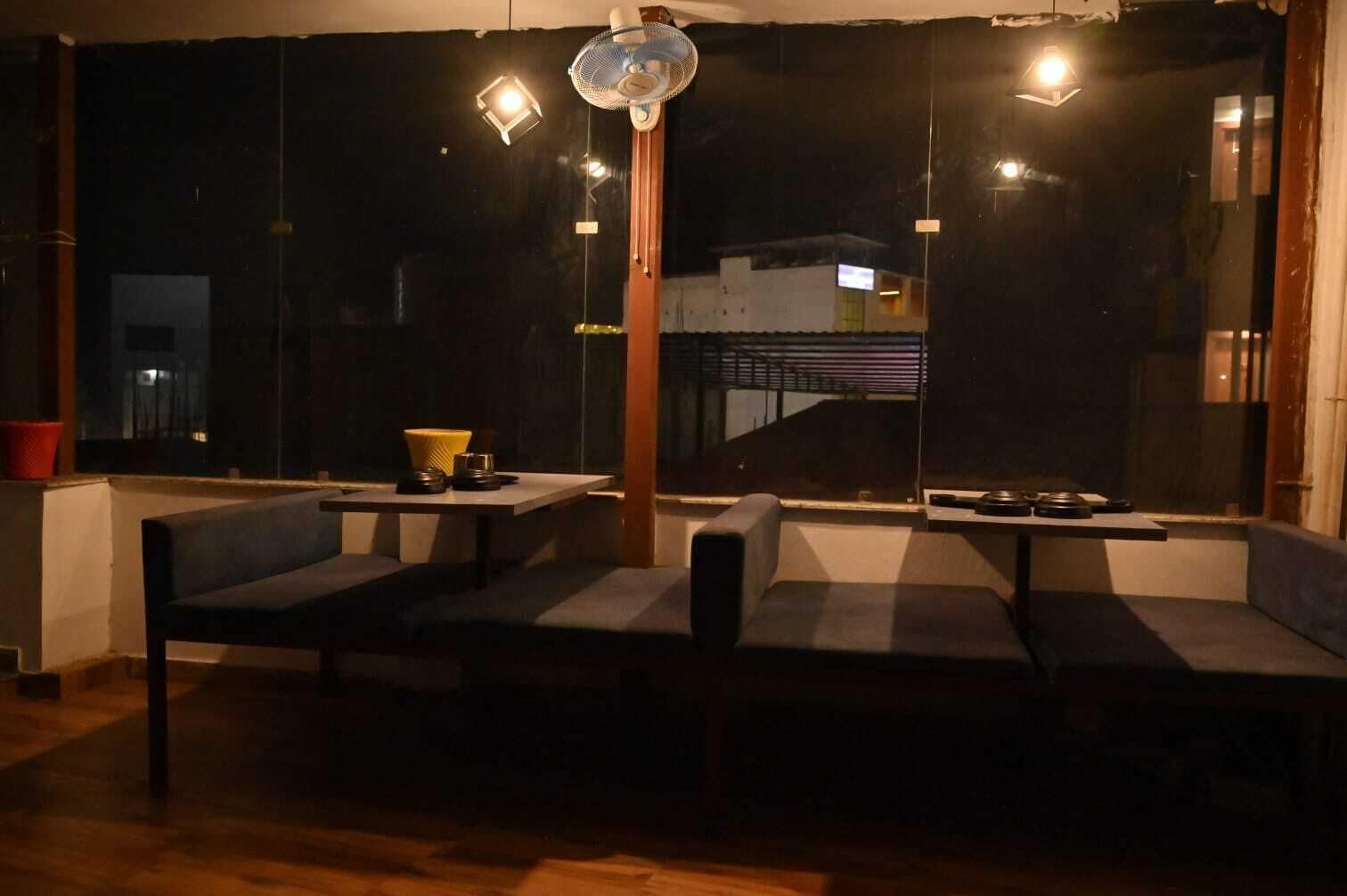 Um quarto com pouca luz, uma janela grande, dois sofás, uma mesinha e um ventilador.