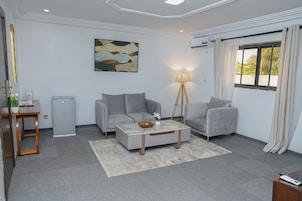 Uma sala de estar moderna com um sofá cinzento, uma mesa de centro e um abajur. Há um quadro na parede e uma janela com cortinas.