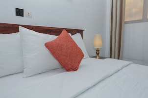 Quarto de hotel com cama, roupa de cama branca, um travesseiro vermelho, abajur e cabeceira de madeira.