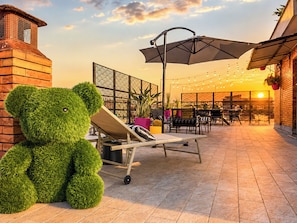 Uma área de pátio com uma grande escultura de urso verde, assentos externos e luzes de corda.