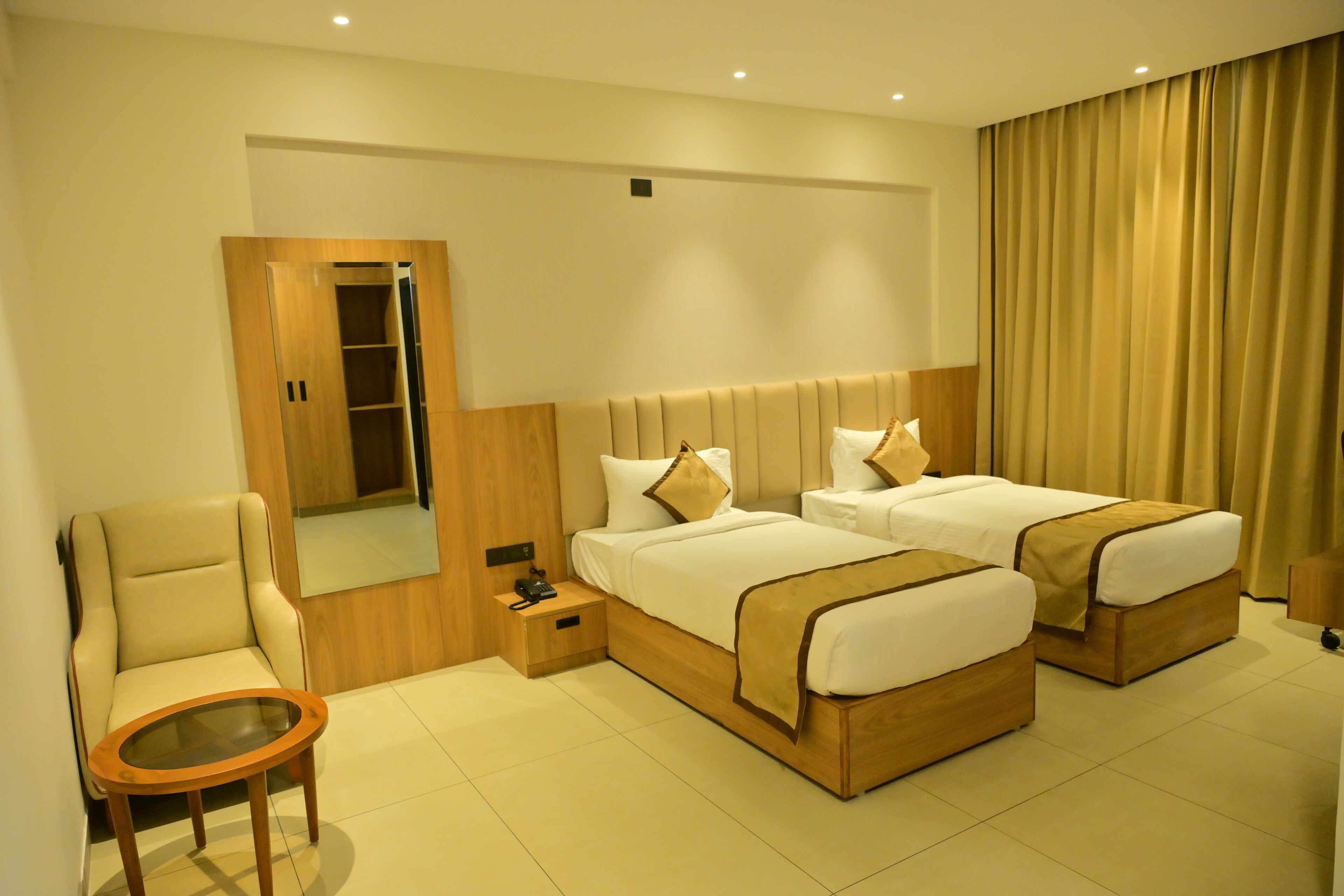 Quarto de hotel com duas camas, uma cadeira, uma mesinha, um criado-mudo e um espelho.