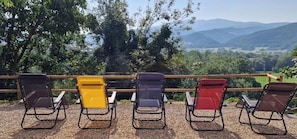 Quatro cadeiras de exterior com estrutura de metal e assentos de tecido, dispostas em fila sobre um piso de cascalho, com uma vedação de madeira e uma vista panorâmica de montanhas e vegetação ao fundo.