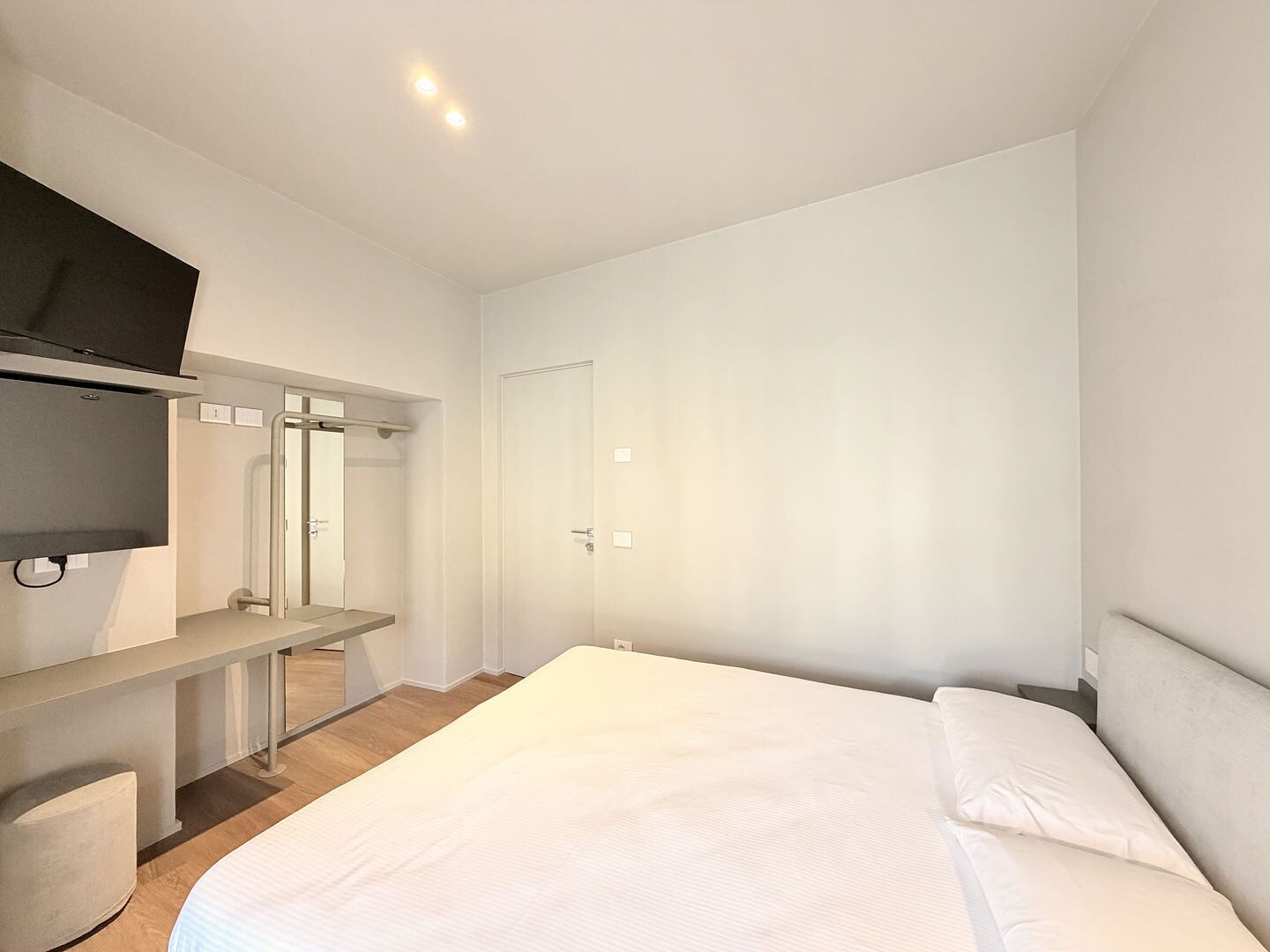 Quarto com cama, televisão na parede, um banco e uma porta.