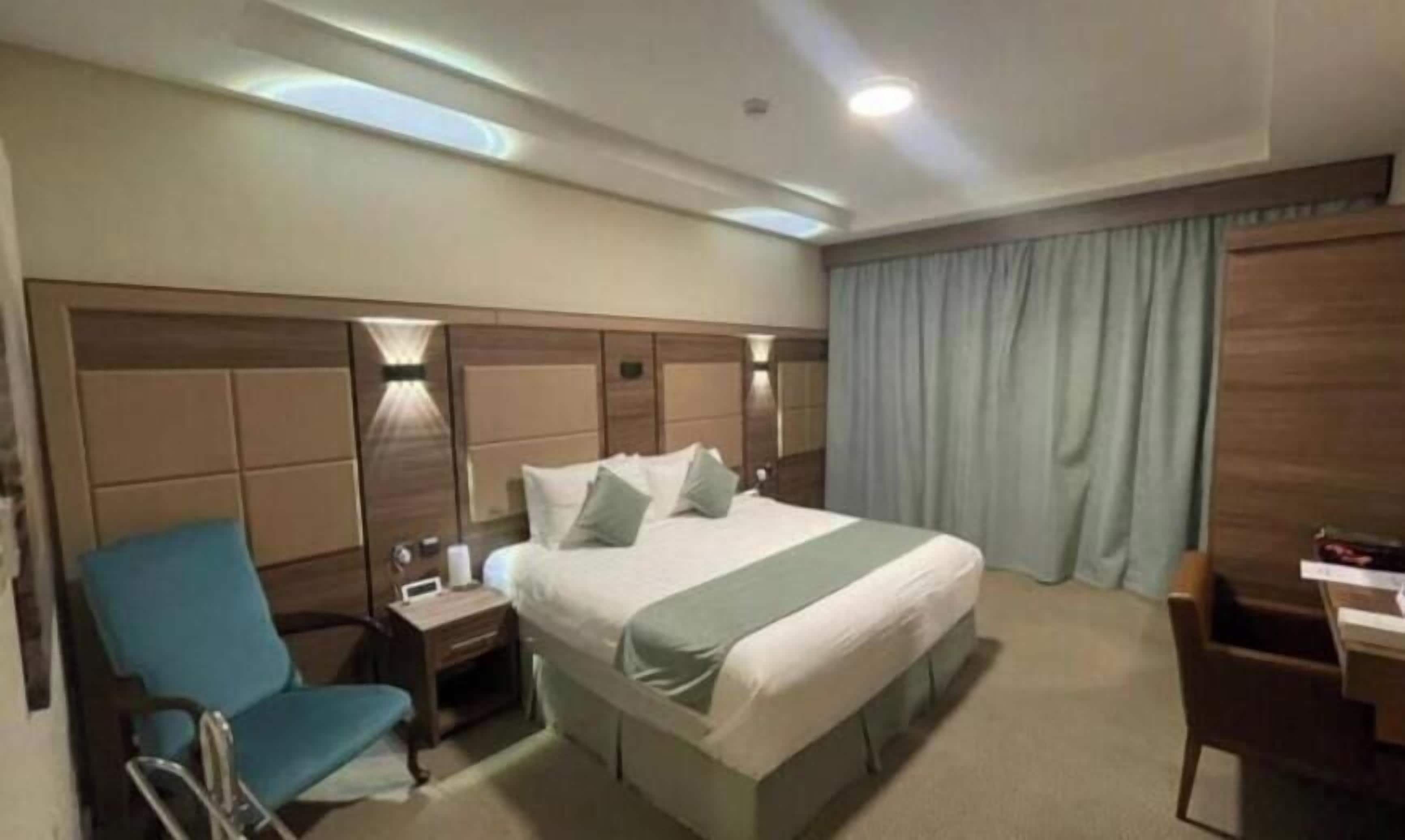 Quarto de hotel com cama, escrivaninha, cadeira e mesa de cabeceira.