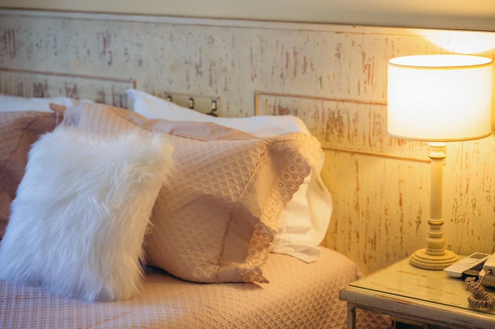 Uma cama com travesseiro de plumas branco e manta texturizada, luminária de cabeceira e telefone em mesa de cabeceira de madeira.