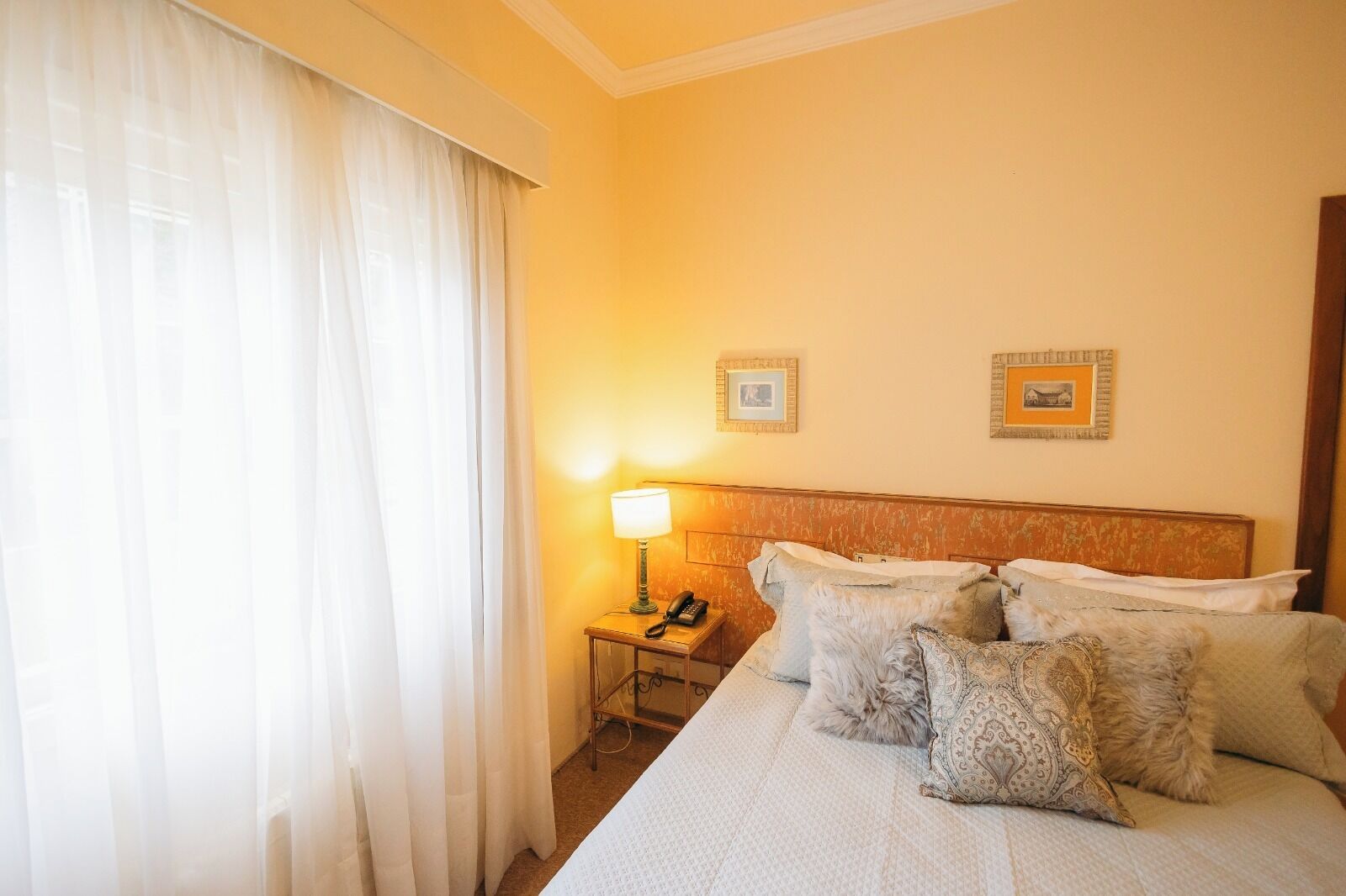 Quarto de hotel com uma cama, uma mesinha de cabeceira com abajur e duas fotos emolduradas na parede.