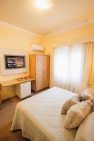 Quarto com cama, escrivaninha com televisão, guarda-roupa e janela com cortinas.