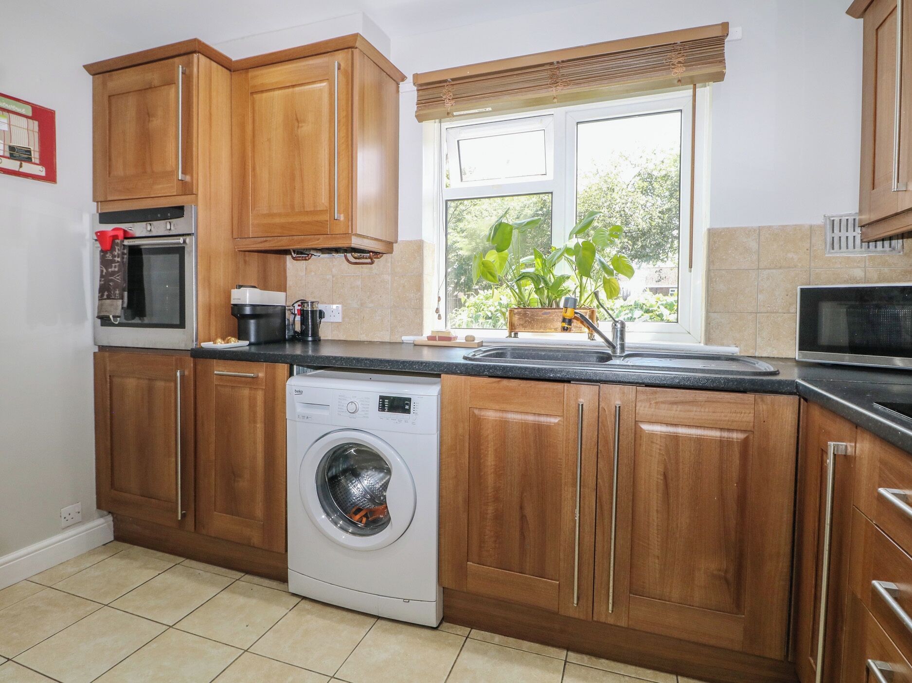 Cozinha com armários de madeira, máquina de lavar, micro-ondas e janela com vista para o verde.