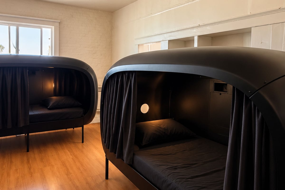 Quarto moderno com duas camas pretas e curvas, cortinas, piso de madeira e janela com luz natural.