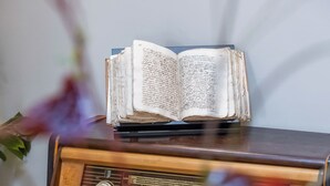 Um rádio antigo com um livro aberto em cima, uma mesa de vidro e um vaso com flores.