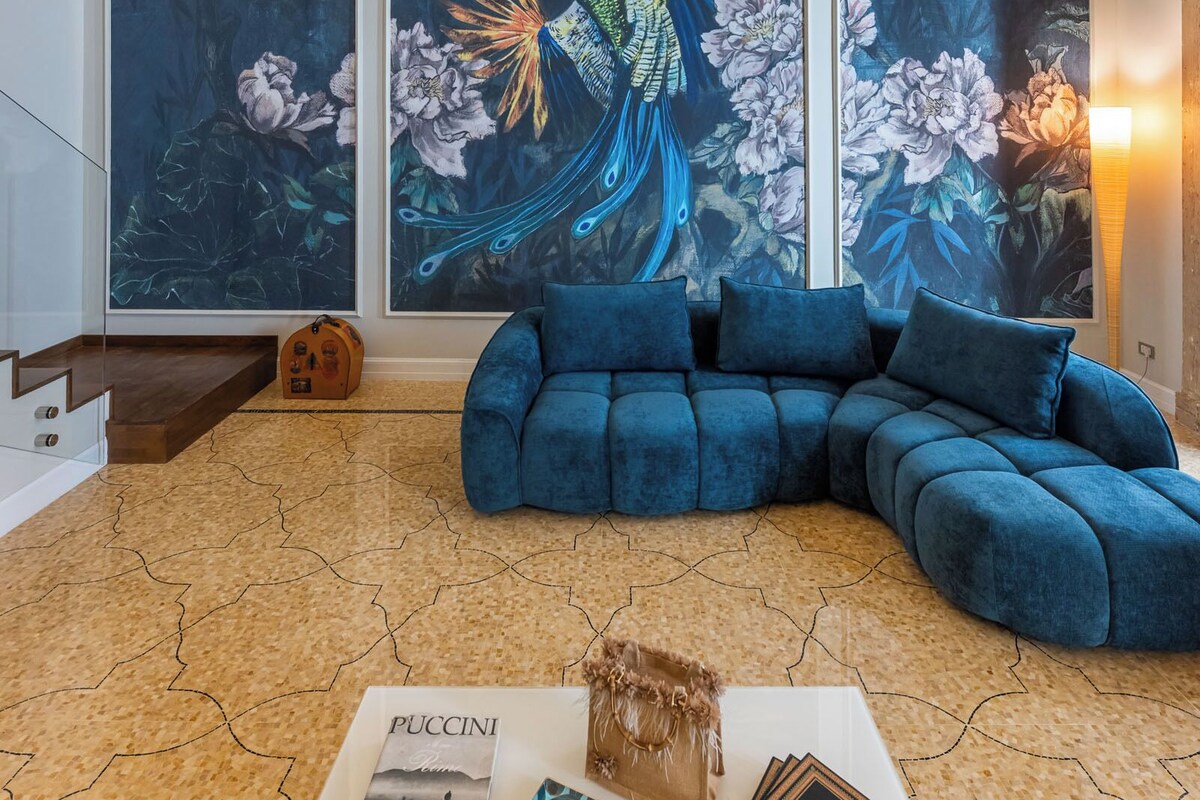 Uma sala de estar moderna com um sofá seccional azul, uma mesa de centro branca com livros e um objeto decorativo, e uma grande janela com um mural de pavão.