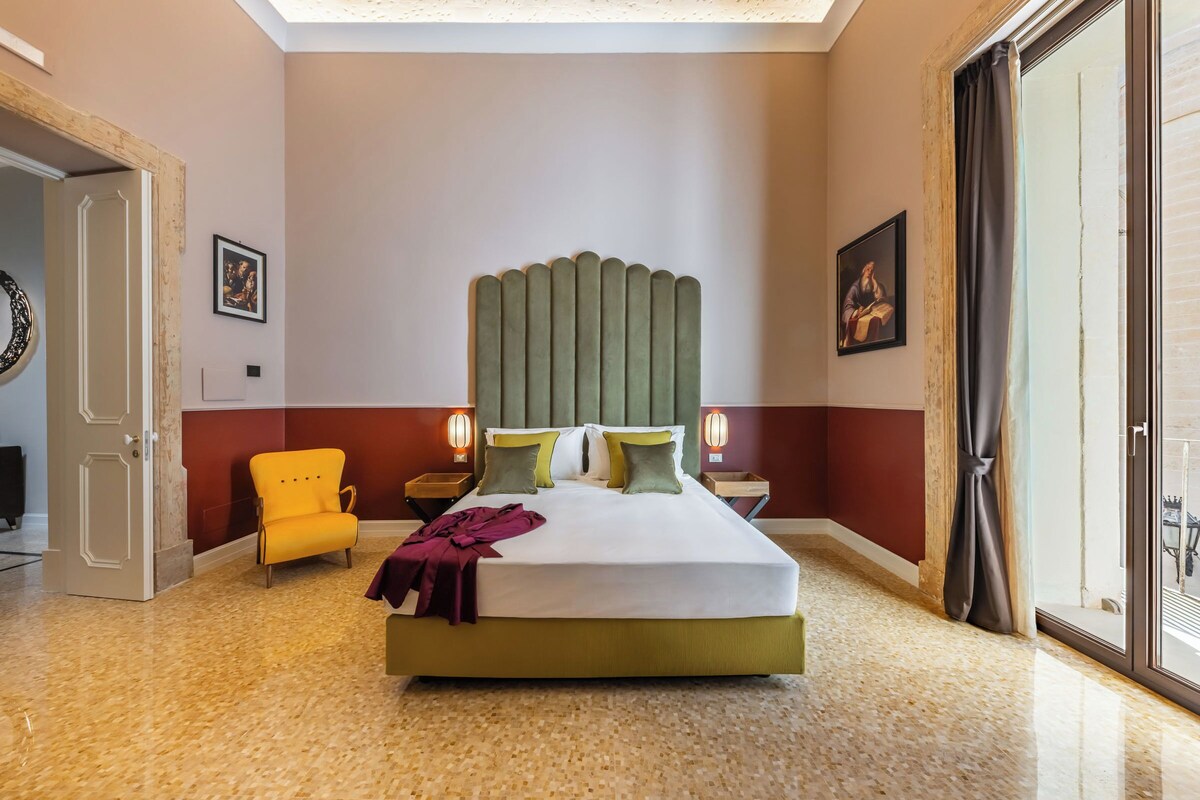 Quarto de hotel com uma cama grande, uma poltrona amarela, duas mesas de cabeceira com luminárias e uma porta que dá para outro quarto.