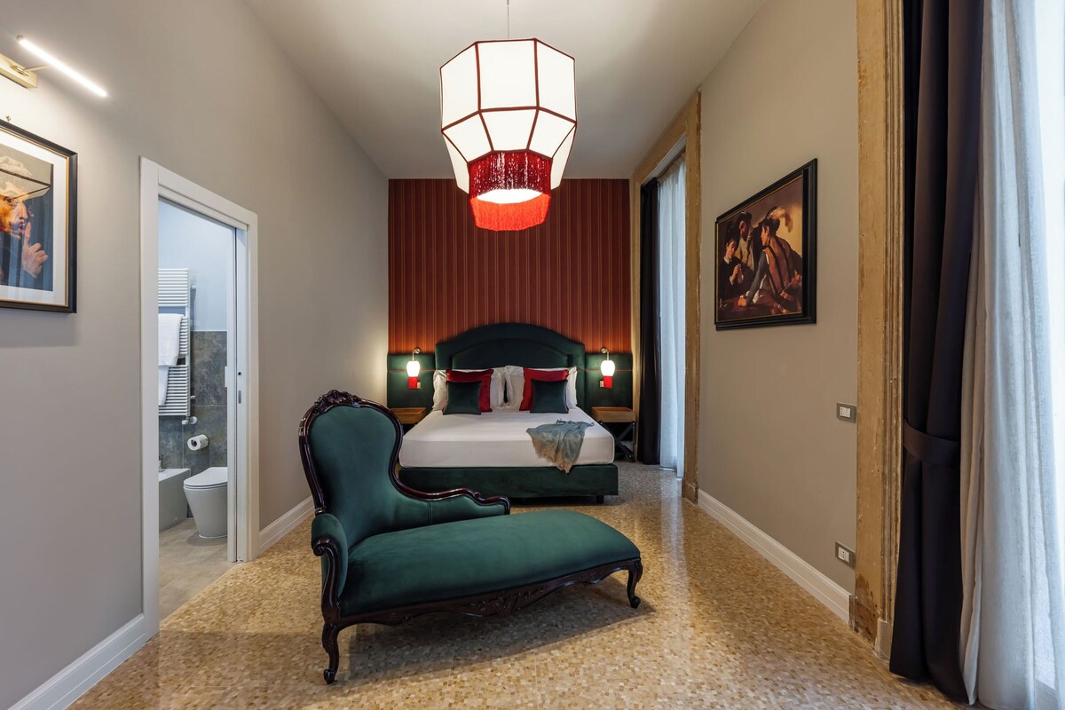 Quarto de hotel com cama, poltrona verde, quadro e corredor que dá para um banheiro.