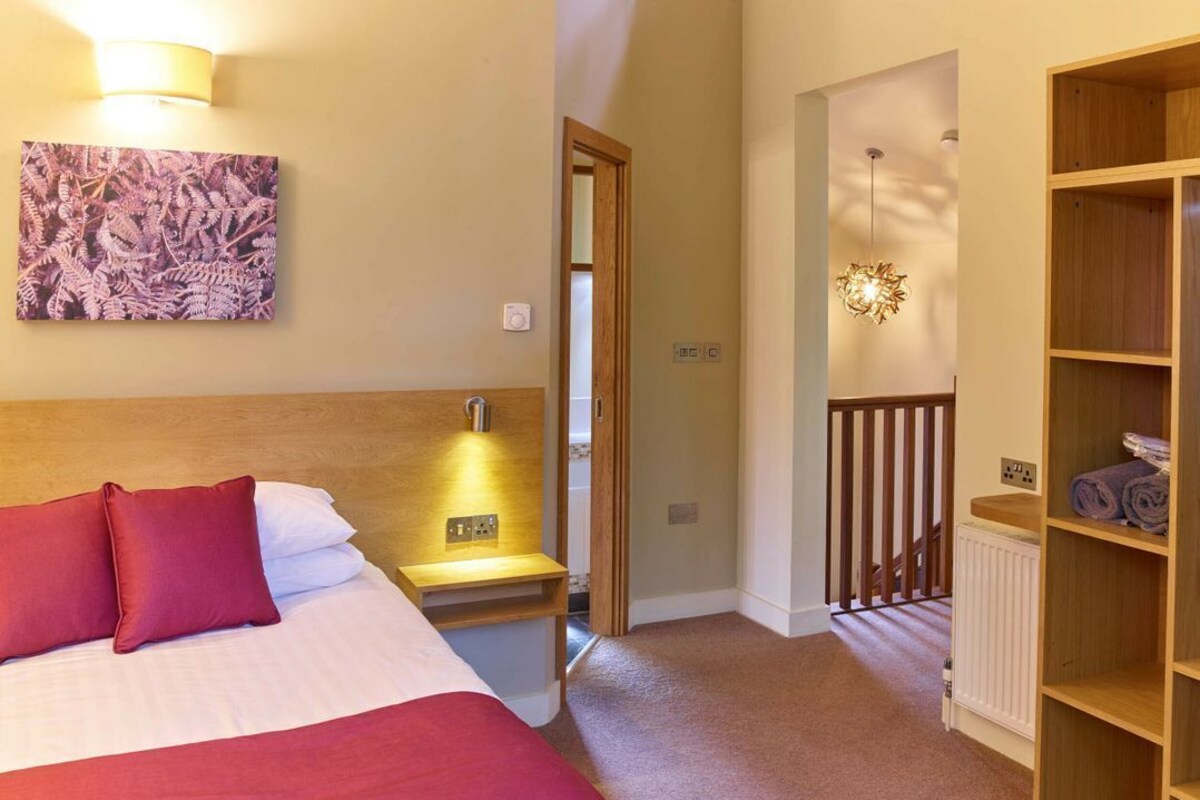 Quarto de hotel com cama, mesa de cabeceira, luminária fixada na parede, cabeceira de madeira, prateleira com toalhas e porta que dá para outro quarto.