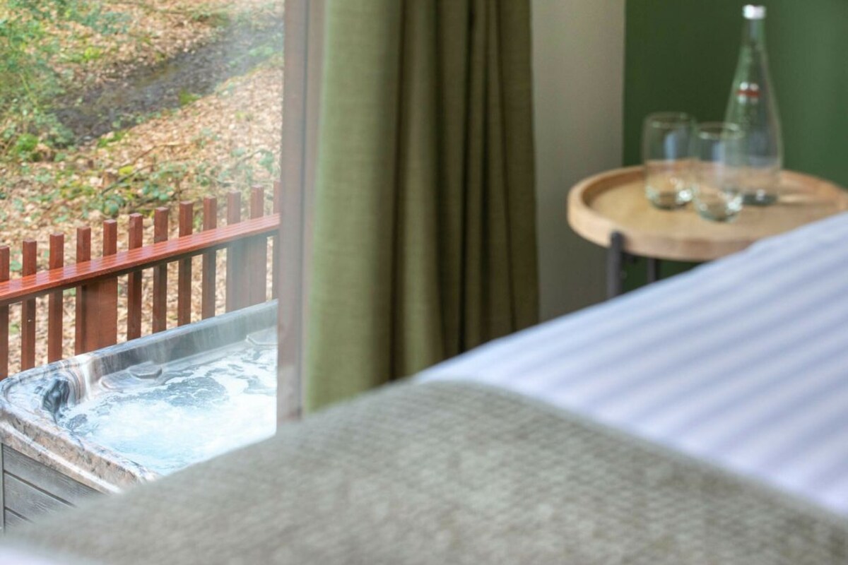 Quarto com cama, janela com vista para um deck de madeira e uma mesinha com copos e uma garrafa.