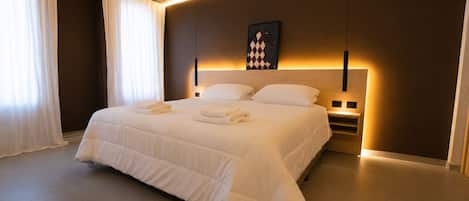 Quarto de hotel com cama grande, roupa de cama branca, cabeceira com iluminação integrada e uma obra de arte emoldurada acima da cama.