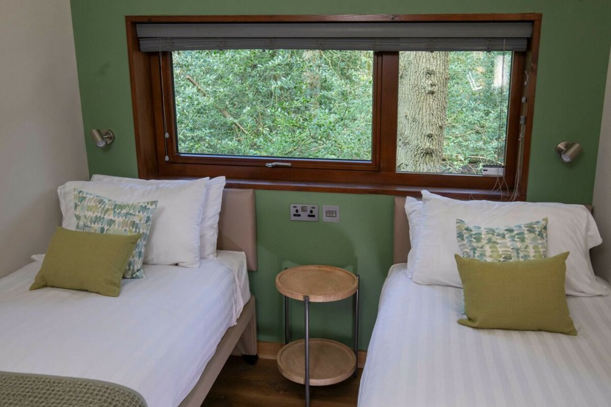 Quarto com duas camas, uma mesa redonda pequena e uma janela com vista para árvores.
