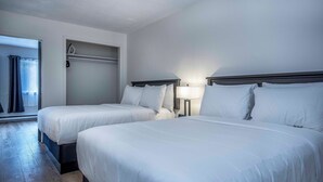 Quarto de hotel com duas camas, um guarda-roupa, uma mesa de cabeceira com abajur e uma janela com cortinas.