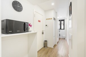 Um corredor com micro-ondas numa prateleira, relógio na parede, porta com aviso, lixeira e janela.