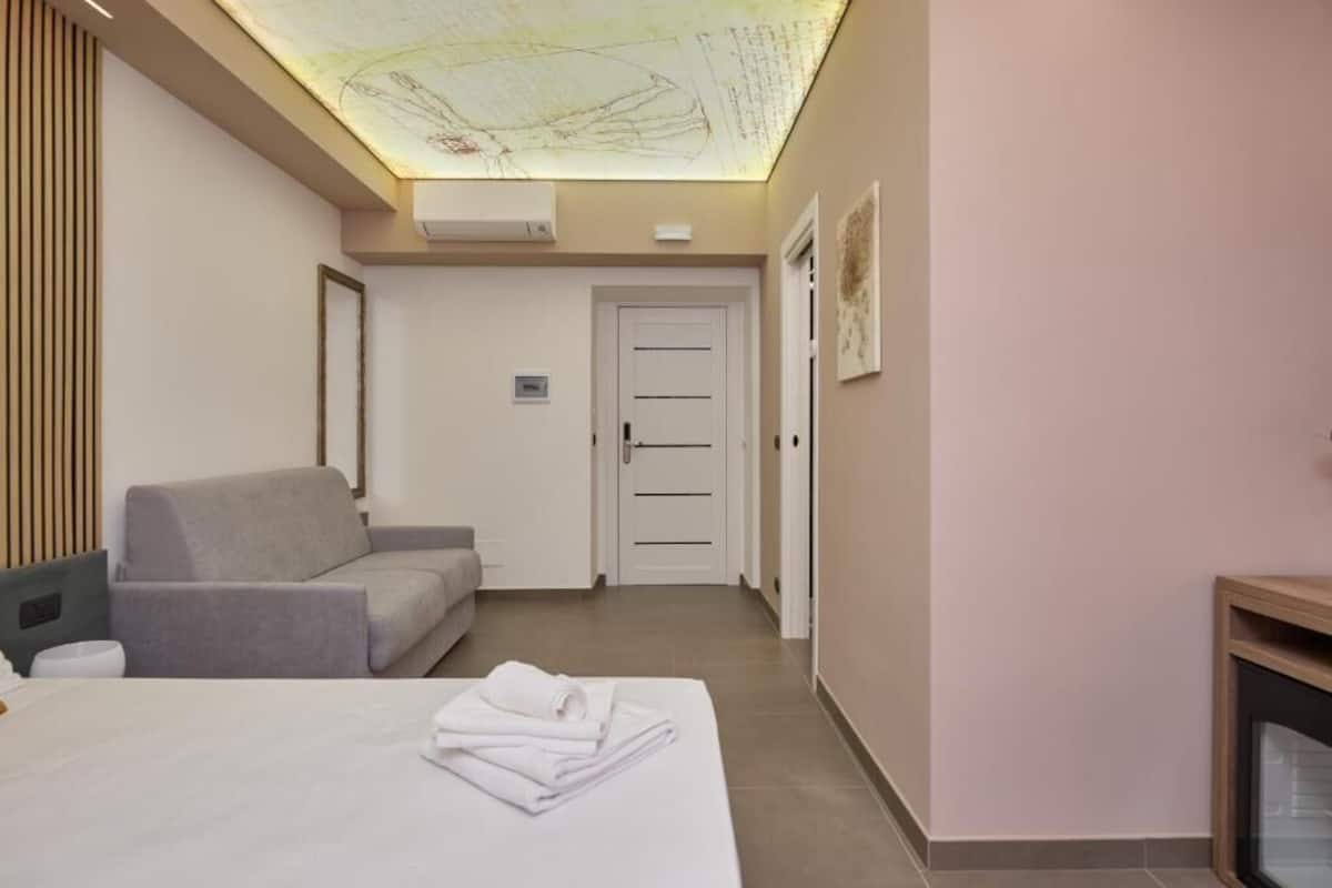 Quarto de hotel com cama, sofá, teto com luminária, porta e ar-condicionado fixado na parede.
