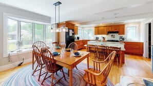 Uma cozinha moderna com armários de madeira, sala de jantar com mesa e cadeiras redondas, e janelas amplas que proporcionam luz natural.