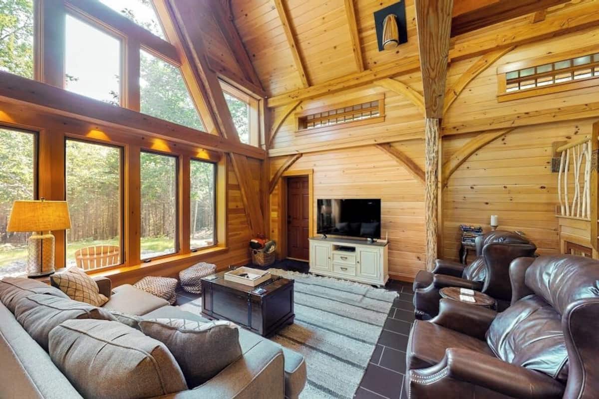 Interior de uma cabana de tronco com uma janela grande, televisão, sofás de couro e lareira.