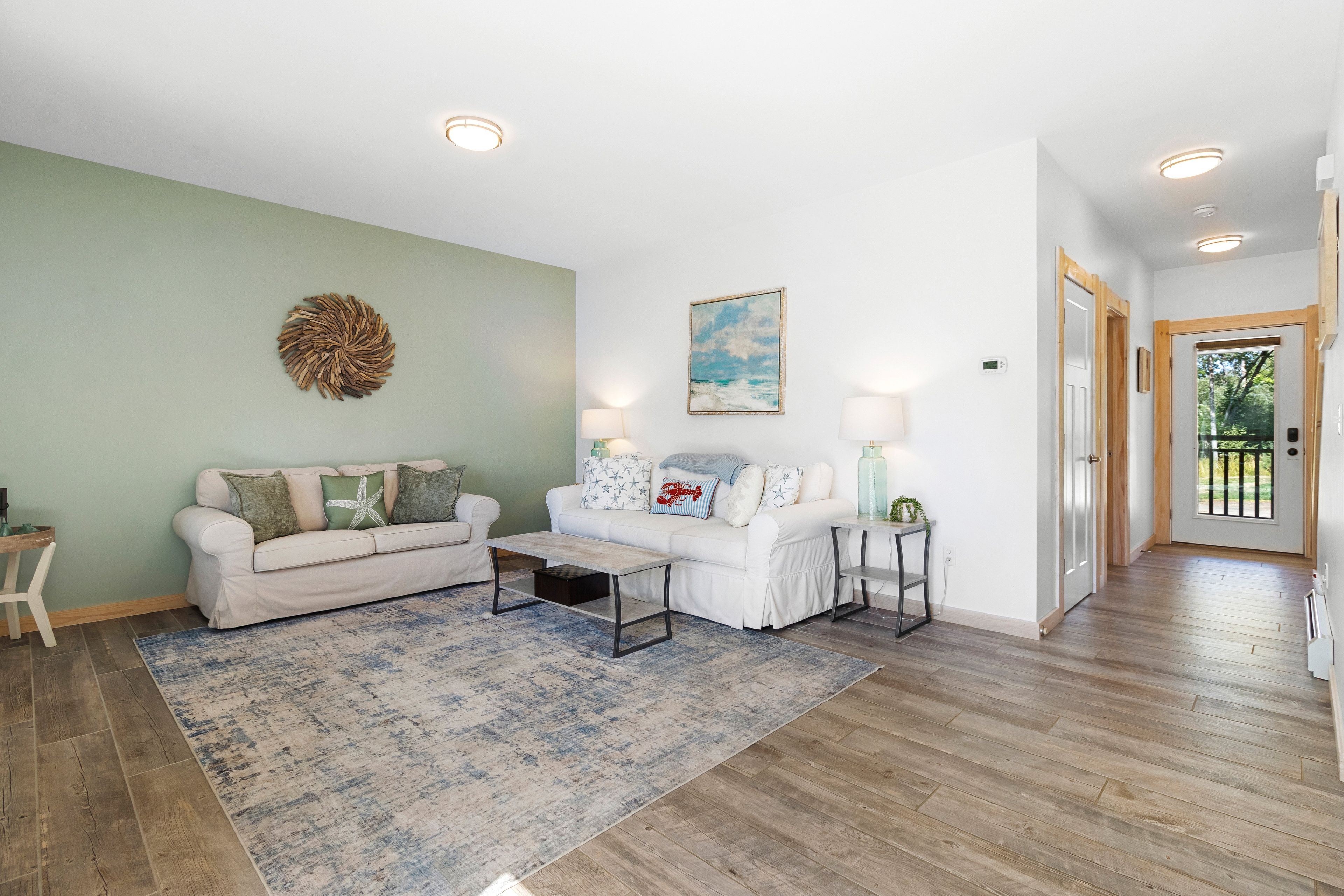 Sala de estar com sofá branco, mesa de centro e tapete. Há um quadro na parede e uma porta que dá para o exterior.