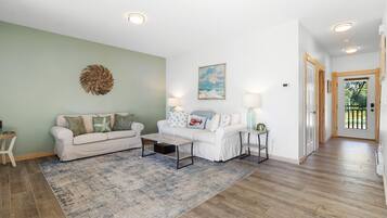 Sala de estar com sofá branco, mesa de centro e tapete. Há um quadro na parede e uma porta que dá para o exterior.