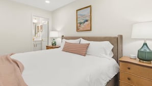 Quarto com uma cama, mesa de cabeceira, luminária, quadro na parede e porta para o banheiro.