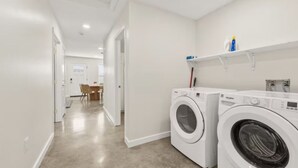 Uma lavanderia com duas máquinas de lavar de abertura frontal, uma área de refeições com cadeiras e um corredor que dá para outro quarto.