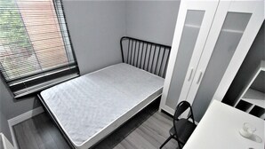 Quarto pequeno com uma cama de solteiro, cabeceira de metal, escrivaninha branca e uma cadeira. Possui janela com persianas, guarda-roupa com portas espelhadas e radiador no chão.