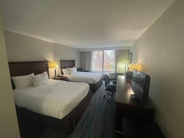 Quarto de hotel com duas camas, uma escrivaninha com um computador e uma janela grande.