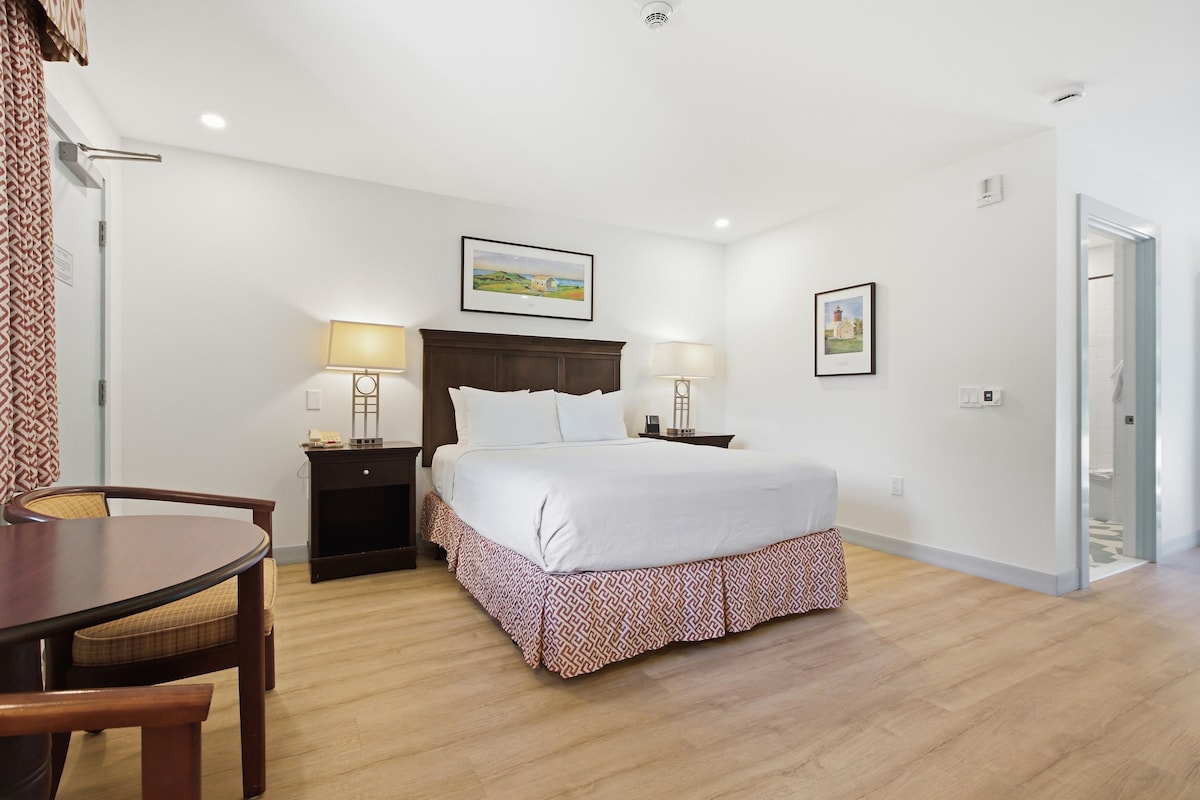Quarto de hotel com cama, mesas de cabeceira, uma cadeira e um quadro na parede.
