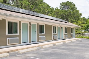 Fileira de quartos de motel com ar condicionado individual, área de estacionamento coberta e paisagem com árvores ao fundo.