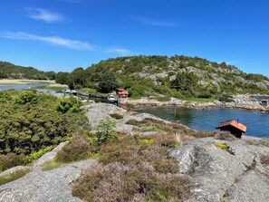Paisagem costeira com um pequeno cais, um edifício com telhado vermelho e uma linha costeira rochosa.