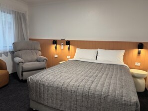 Quarto de hotel com uma cama grande, uma poltrona cinza, uma mesinha de cabeceira redonda e uma luminária fixada na parede.